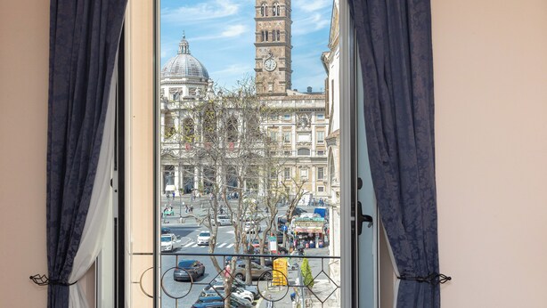 Vista da janela com cortinas, mostrando um edifício histórico com torre do relógio e cúpula, uma rua movimentada e carros estacionados.