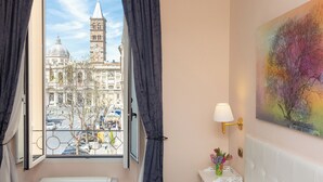 Quarto de hotel com uma grande janela, vista para um edifício histórico, cama, mesa de cabeceira com abajur e um quadro na parede.