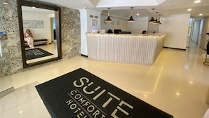 Uma área de recepção de hotel com parede de mármore, balcão de recepção e tapete preto com a inscrição 'SUITE COMFORT HOTEL'.