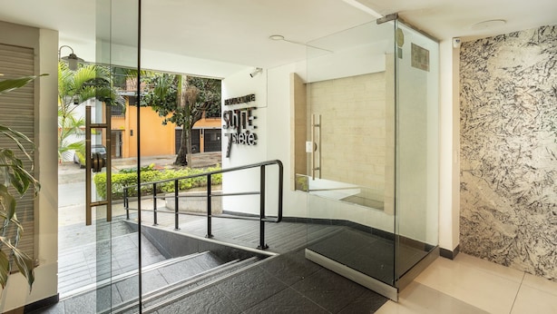 Um saguão de hotel moderno com entrada de vidro, uma escada que leva ao andar superior e vista para a área externa, incluindo uma palmeira e carros estacionados.