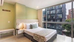 Quarto de hotel moderno com uma cama grande, mesas de cabeceira, um banco e vista para a cidade através de amplas janelas.