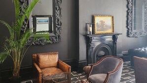 Quarto com paredes escuras, lareira, duas poltronas, um espelho grande e um vaso de planta.