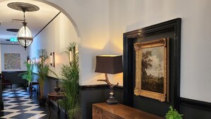 Um hall com piso xadrez, um console de madeira com abajur, um quadro emoldurado e um lustre.