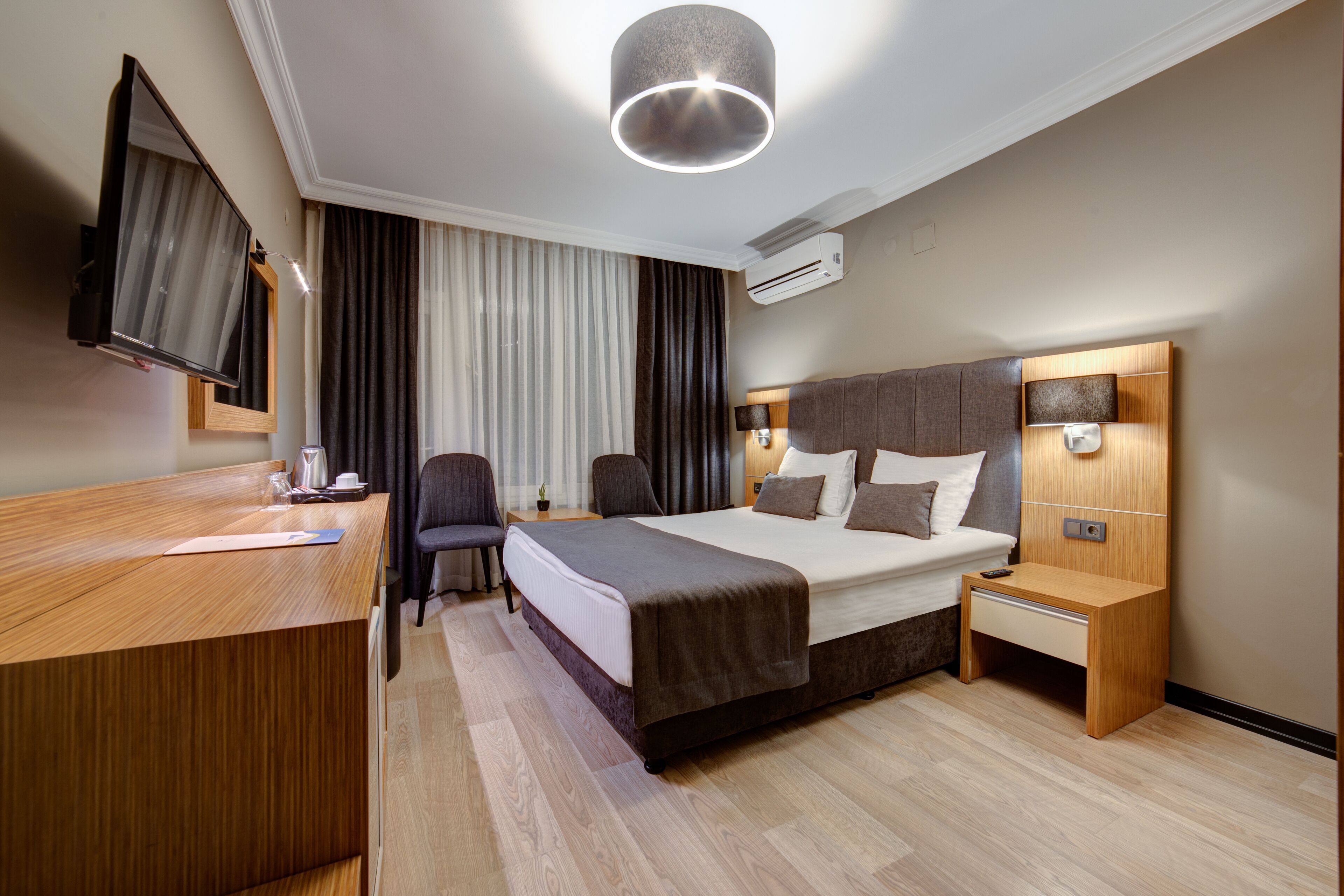 Quarto de hotel moderno com escrivaninha de madeira, cama com cabeceira, mesa de cabeceira, televisão presa na parede e luminária no teto.