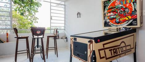 Quarto com mesa de futebol de mesa, um banquinho de bar e uma grande janela com vista para árvores.