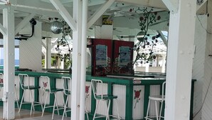 Uma área de estar coberta ao ar livre, com cadeiras brancas e verdes, uma máquina de venda automática da Coca-Cola e uma bola de espelhos.