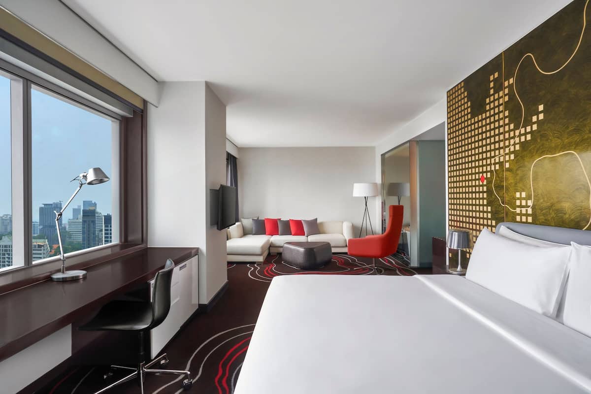 Quarto de hotel com uma cama grande, uma escrivaninha com cadeira, um sofá, uma poltrona vermelha, uma televisão e vista para a cidade.