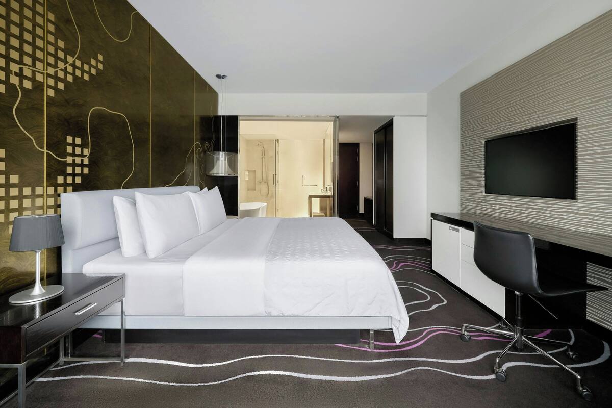 Quarto de hotel moderno com uma cama grande, uma escrivaninha, uma cadeira, uma TV e um banheiro visível através de uma porta aberta.