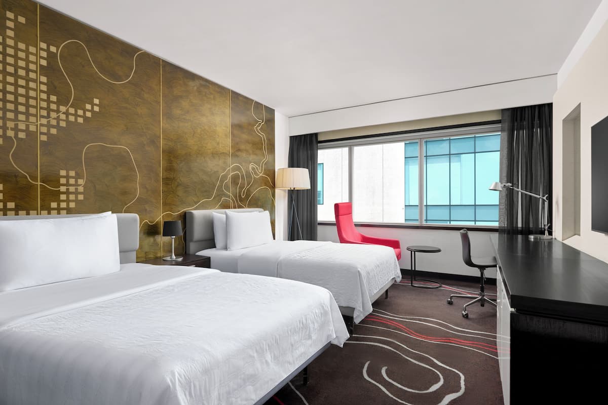 Quarto de hotel com duas camas, uma escrivaninha, uma cadeira vermelha e uma janela grande.