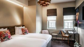 Quarto de hotel com cama grande, escrivaninha, cadeira, mesinha e janela com persianas.