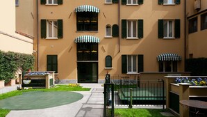 Um edifício de vários andares com entrada central, persianas verdes e um pequeno jardim com quadra de tênis.