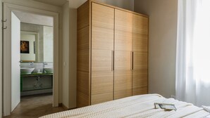 Quarto com um guarda-roupa de madeira grande, uma cama com cobertor branco e um banheiro visível através de uma porta aberta.