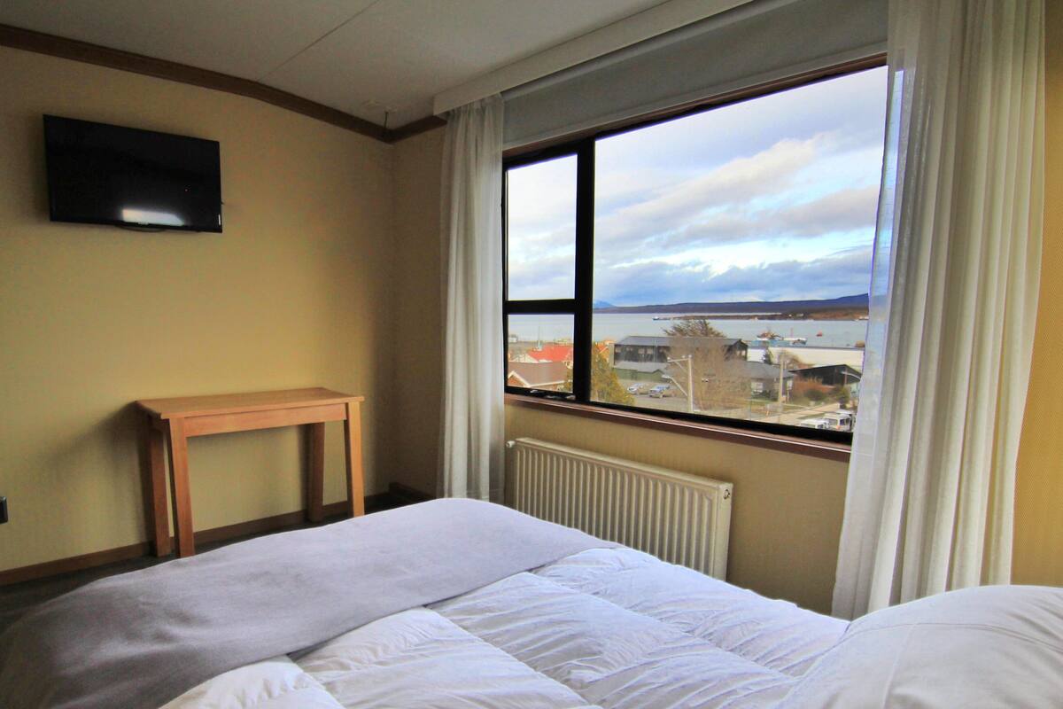 Quarto de hotel com cama, uma pequena mesa de madeira, televisão fixada na parede e janela com vista para um corpo de água e edifícios.