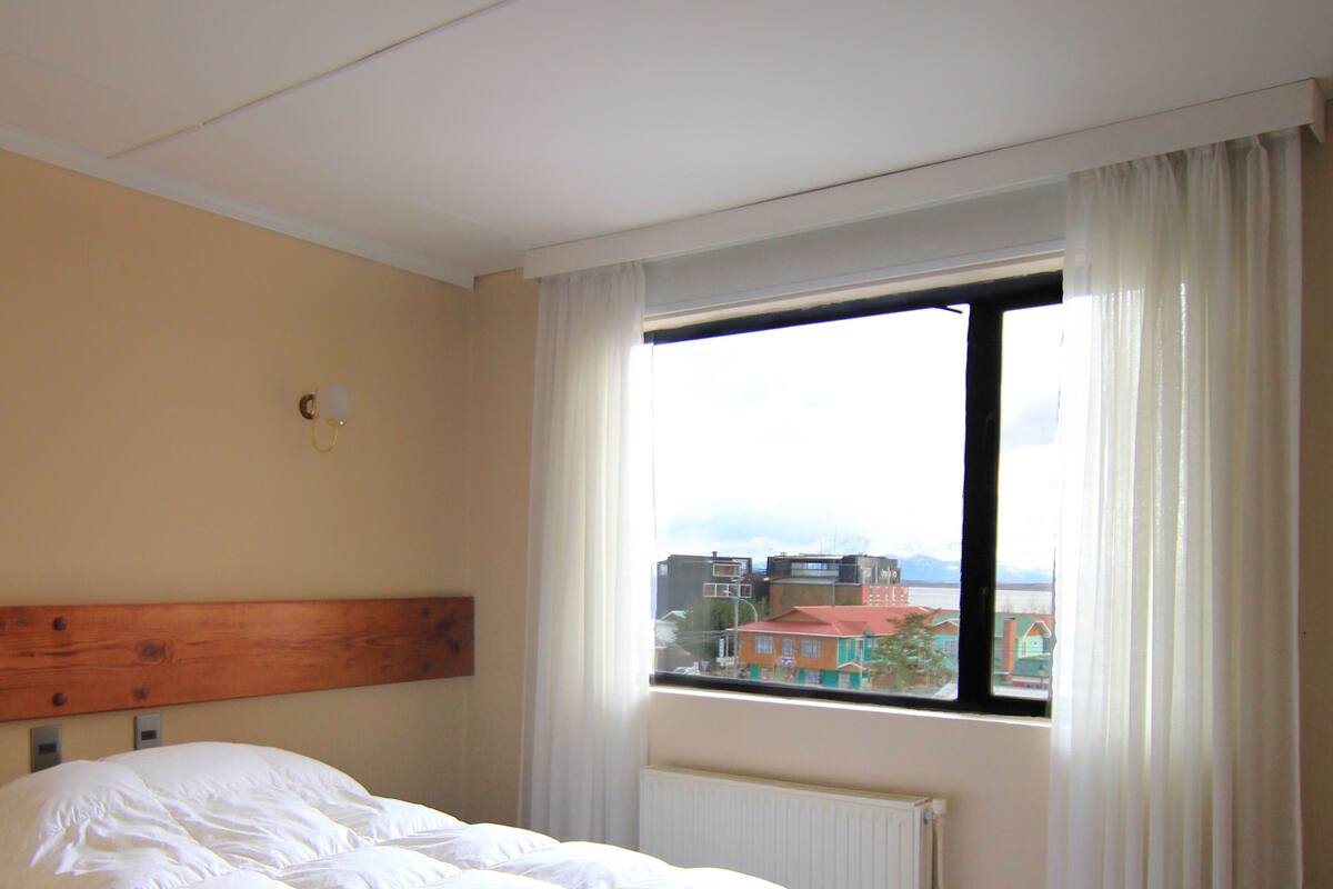Quarto de hotel com uma cama grande, cabeceira de madeira, janela com cortinas e vista para prédios.