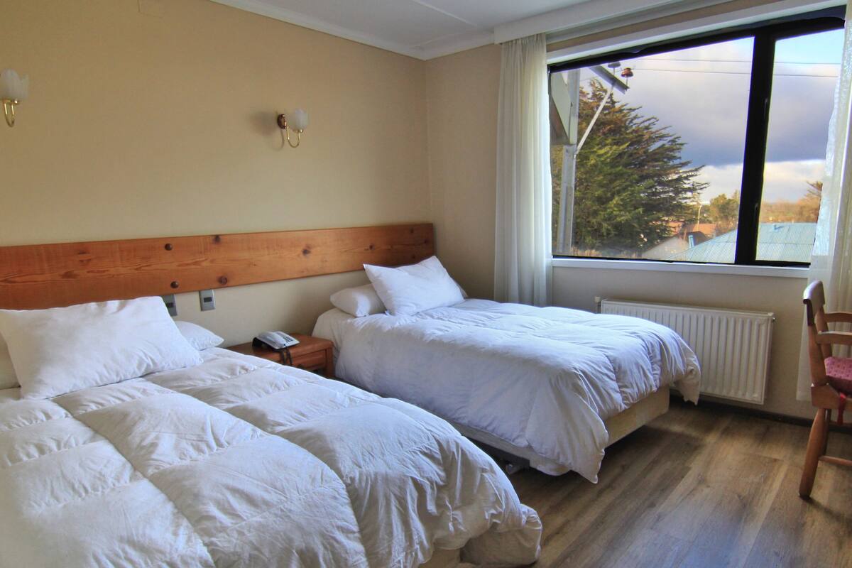 Quarto de hotel com duas camas, uma cabeceira de madeira, uma mesa de cabeceira, uma cadeira e uma janela com vista para árvores e um edifício.