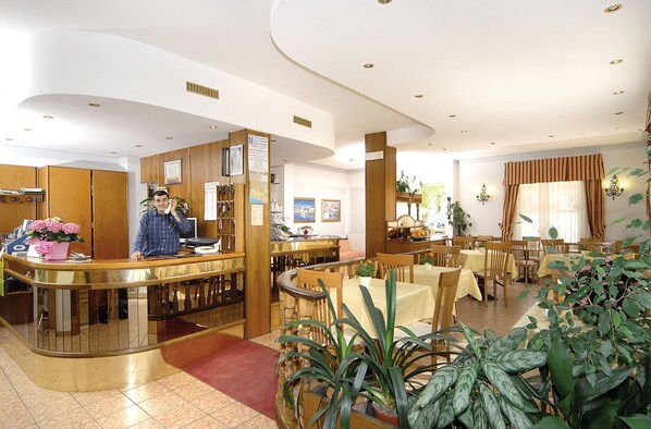 Um saguão de hotel com balcão de recepção, área de refeições e uma grande planta em vaso.