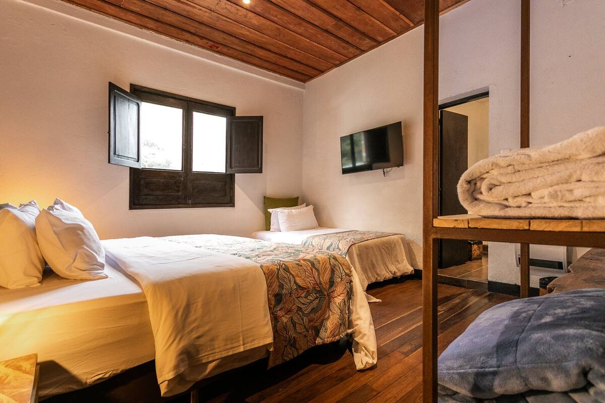 Quarto com duas camas, teto de madeira, TV na parede e janela com persianas.