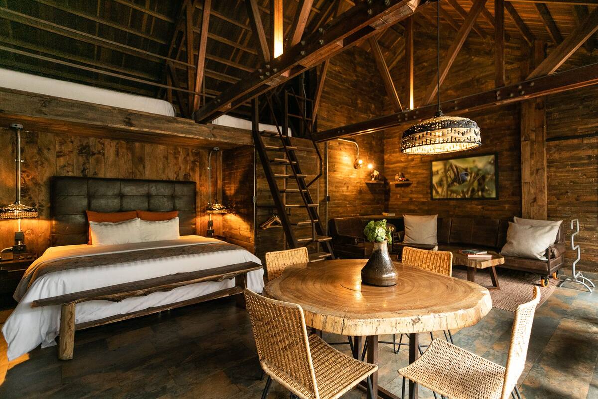 Um quarto rústico de madeira com uma cama, uma mesa redonda de madeira com cadeiras, um sofá e uma escada que dá acesso a um mezanino.