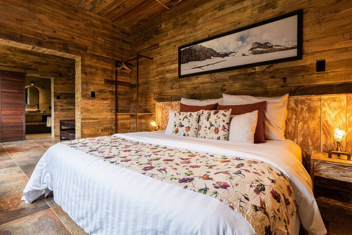 Quarto com painéis de madeira, uma cama grande, um quadro na parede e um banco perto da porta.