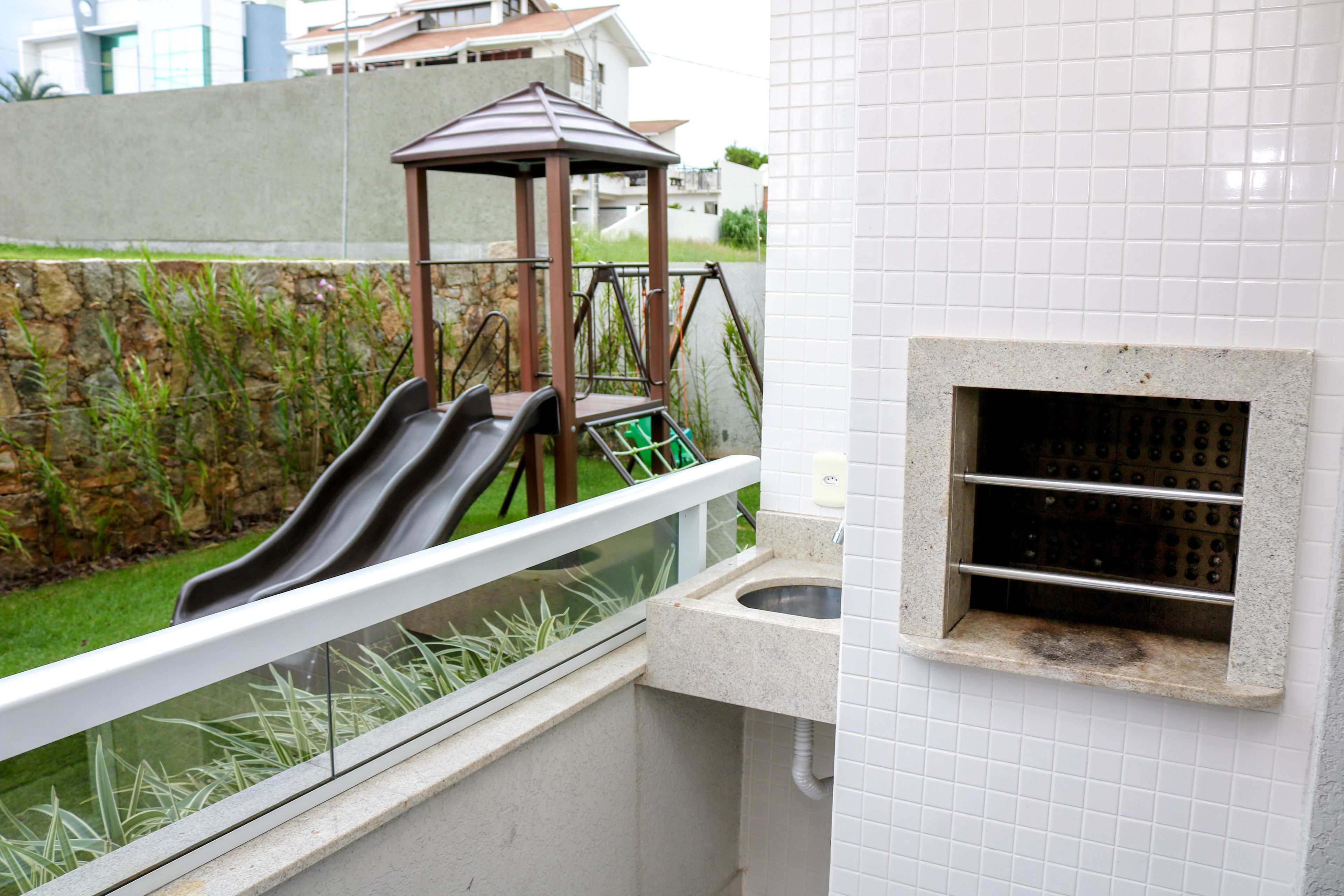 Varanda residencial com área de recreação infantil, incluindo escorrega e balanço, adjacente a uma parede de azulejos brancos e uma janela.