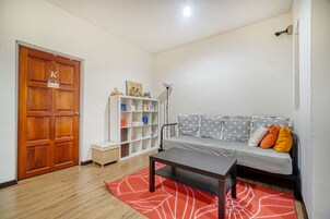 Uma sala de estar com porta de madeira, estante de livros, sofá com almofadas e um tapete vermelho.