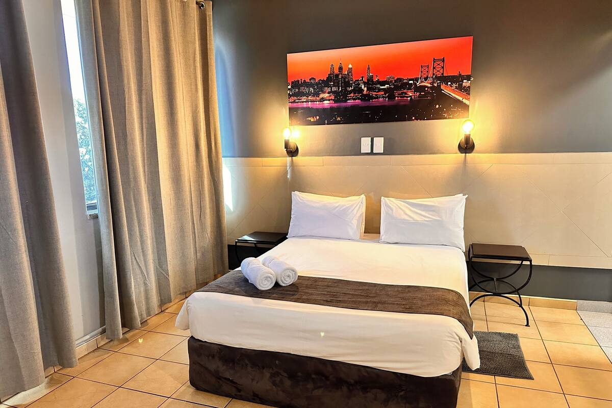 Quarto de hotel com uma cama grande, duas mesas de cabeceira, uma cabeceira com uma pintura de paisagem urbana e vista para a cidade pela janela.