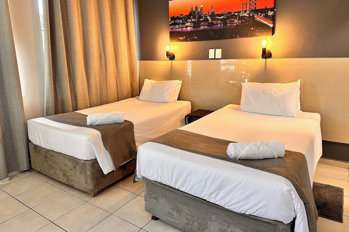 Quarto de hotel com duas camas, cabeceira, pintura de um horizonte urbano e mesa de cabeceira.