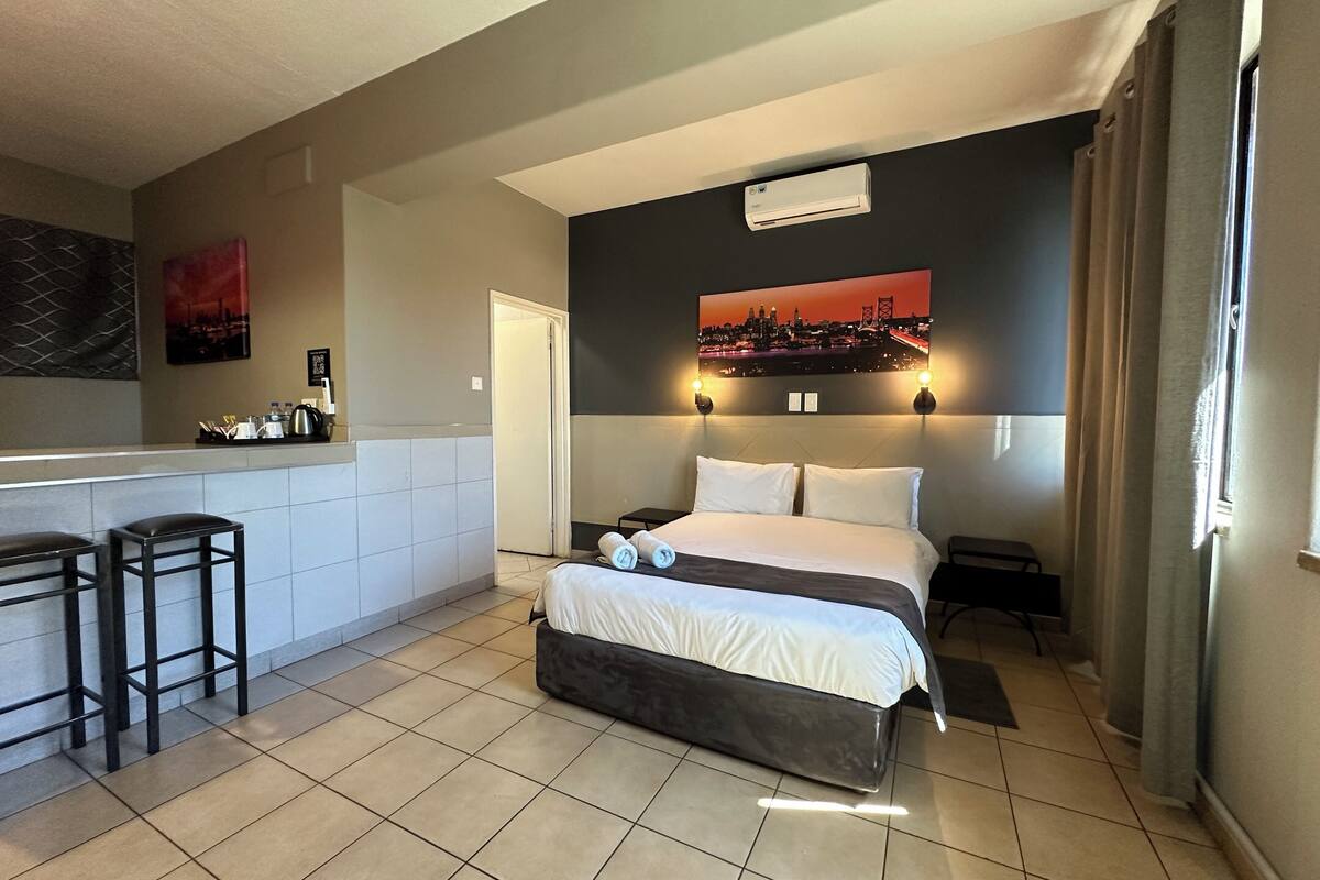 Quarto de hotel com uma cama, uma pequena mesa com cadeira, um quadro na parede e um aparelho de ar condicionado.