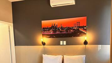 Quarto de hotel com cama, pintura de paisagem urbana, ar condicionado e duas luminárias fixadas na parede.