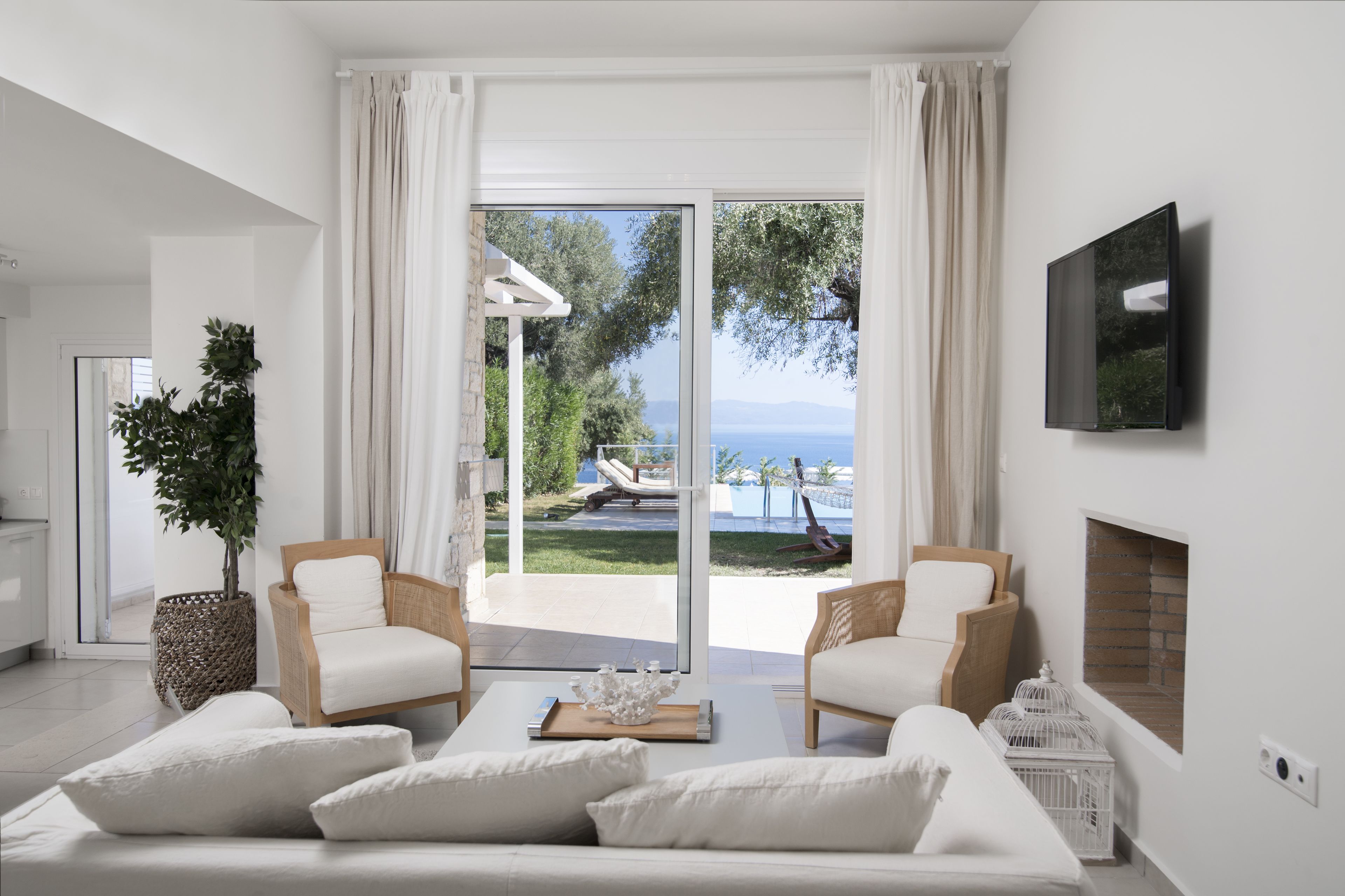 Uma sala de estar moderna com um sofá branco, duas poltronas, uma mesa de centro e uma grande porta de vidro deslizante que dá para uma varanda com vista para uma piscina e áreas verdes.