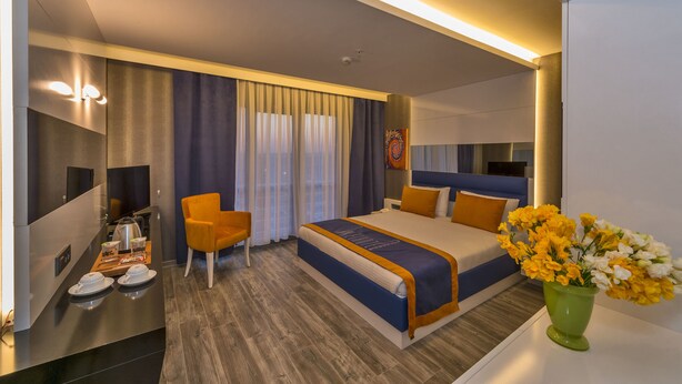 Quarto de hotel moderno com cama, escrivaninha com cadeira, pequena mesa com bandeja de itens de café da manhã e um vaso de flores.