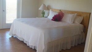 Cama bem arrumada com colcha branca e uma flor rosa decorativa. Um criado-mudo com uma luminária e uma pequena planta ao lado da cama.