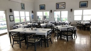 Uma sala de jantar com várias mesas e cadeiras, janelas amplas e obras de arte emolduradas nas paredes.