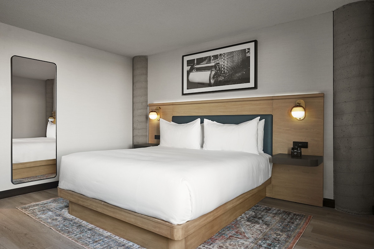 Quarto de hotel moderno com uma cama grande, uma mesinha de cabeceira, uma luminária de parede, um quadro emoldurado e um espelho.