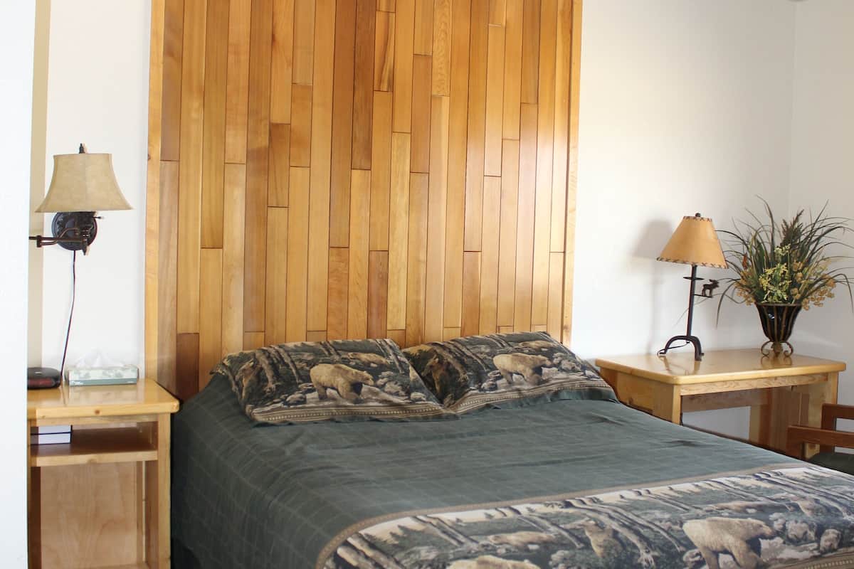Quarto com cabeceira de madeira, cama com colcha verde, duas mesas de cabeceira com abajures e uma pequena escrivaninha de madeira com uma planta.