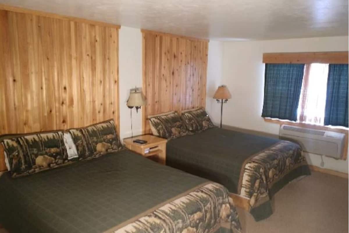 Quarto com duas camas, painéis de madeira nas paredes, janela com cortinas e abajur em uma mesa de cabeceira.