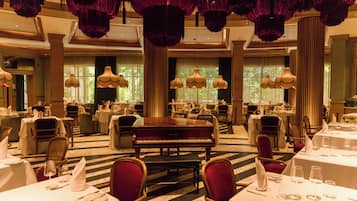 Uma sala de jantar formal, com mesas postas para o serviço, um piano de cauda e lustres elegantes.