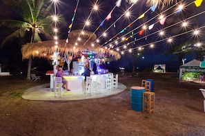 Uma área de bar ao ar livre com telhado de palha, banquetas brancas e um balcão.