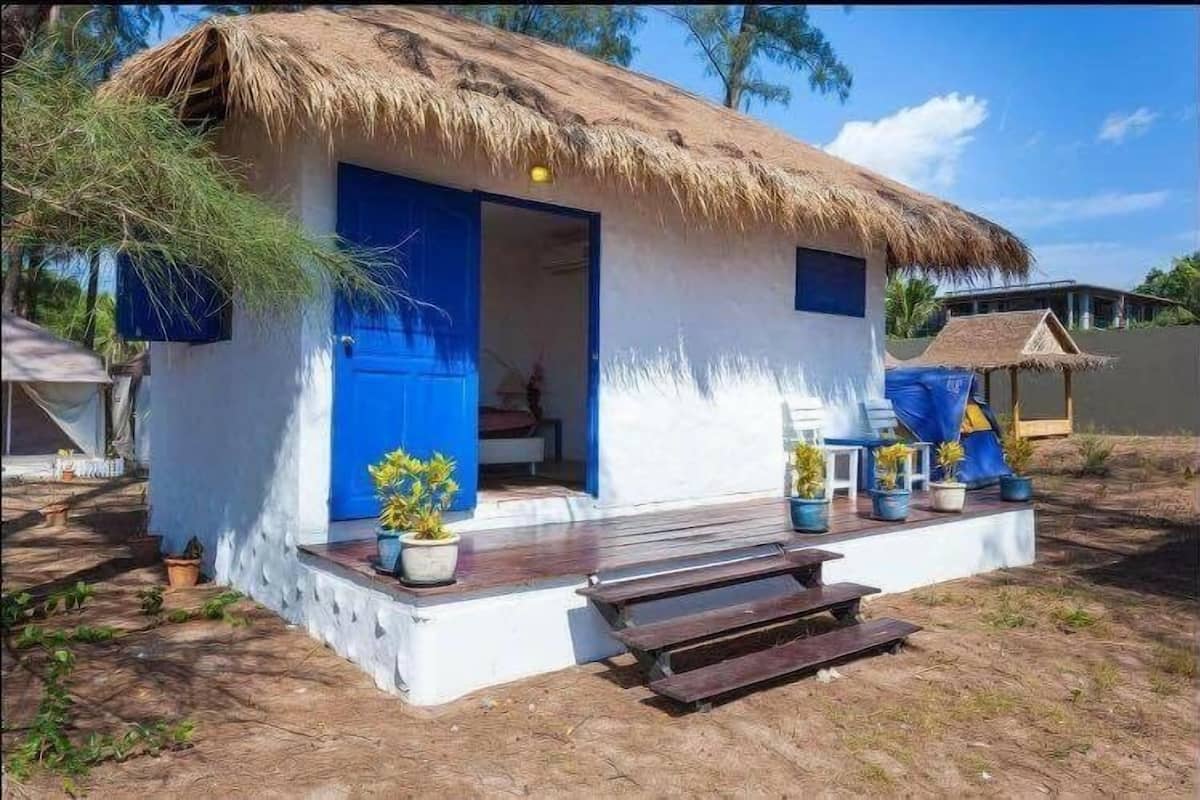 Uma pequena casa térrea com telhado de palha, porta azul e varanda com plantas em vasos.