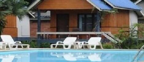 Um bangalô de madeira com piscina, cadeiras de lounge brancas e telhado azul.