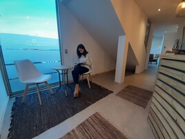 Um interior moderno com uma mulher sentada numa cadeira perto de uma janela grande, uma mesa pequena e um armário de madeira.