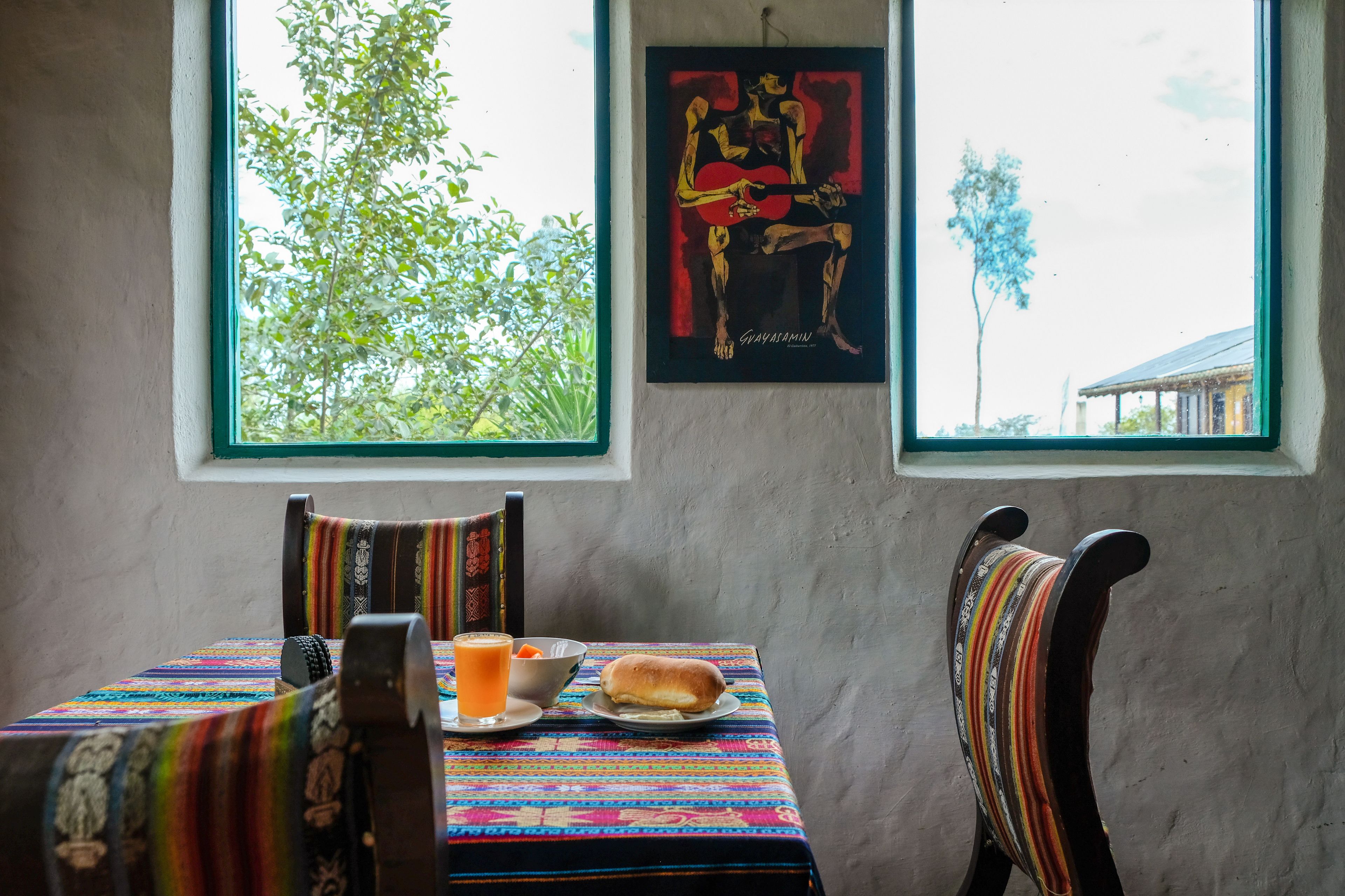 Uma área de refeições com uma mesa posta para dois, com toalha de mesa colorida e um prato com sanduíche e copo de suco de laranja.