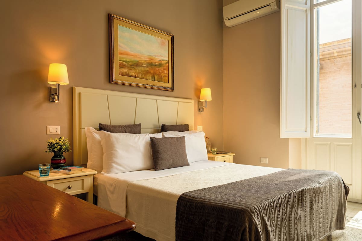 Quarto de hotel com cama, abajures, um quadro na parede e uma janela.