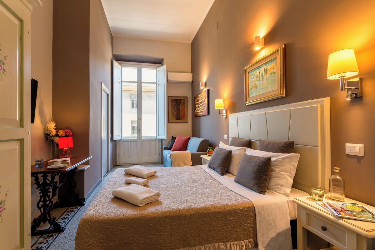 Quarto de hotel com cama grande, mesas de cabeceira, escrivaninha com luminária e janela com vista.