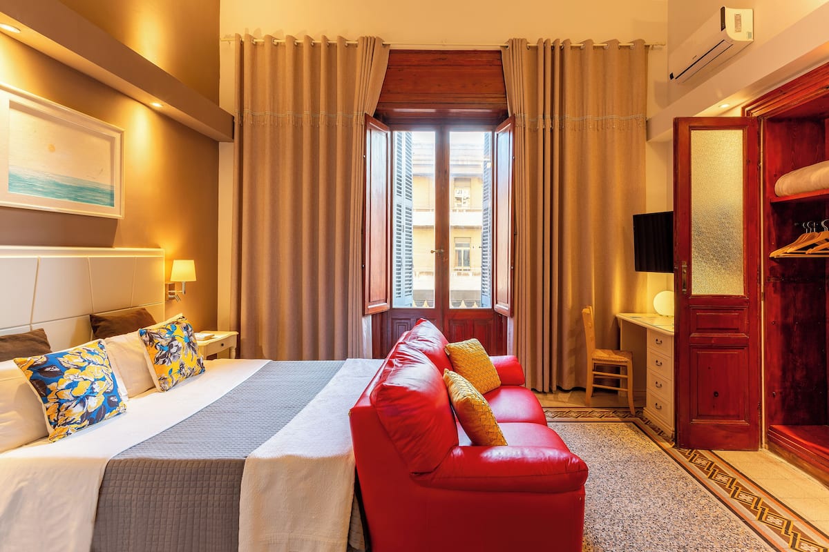 Quarto de hotel com uma cama grande, um sofá vermelho, uma escrivaninha com cadeira e um guarda-roupa.