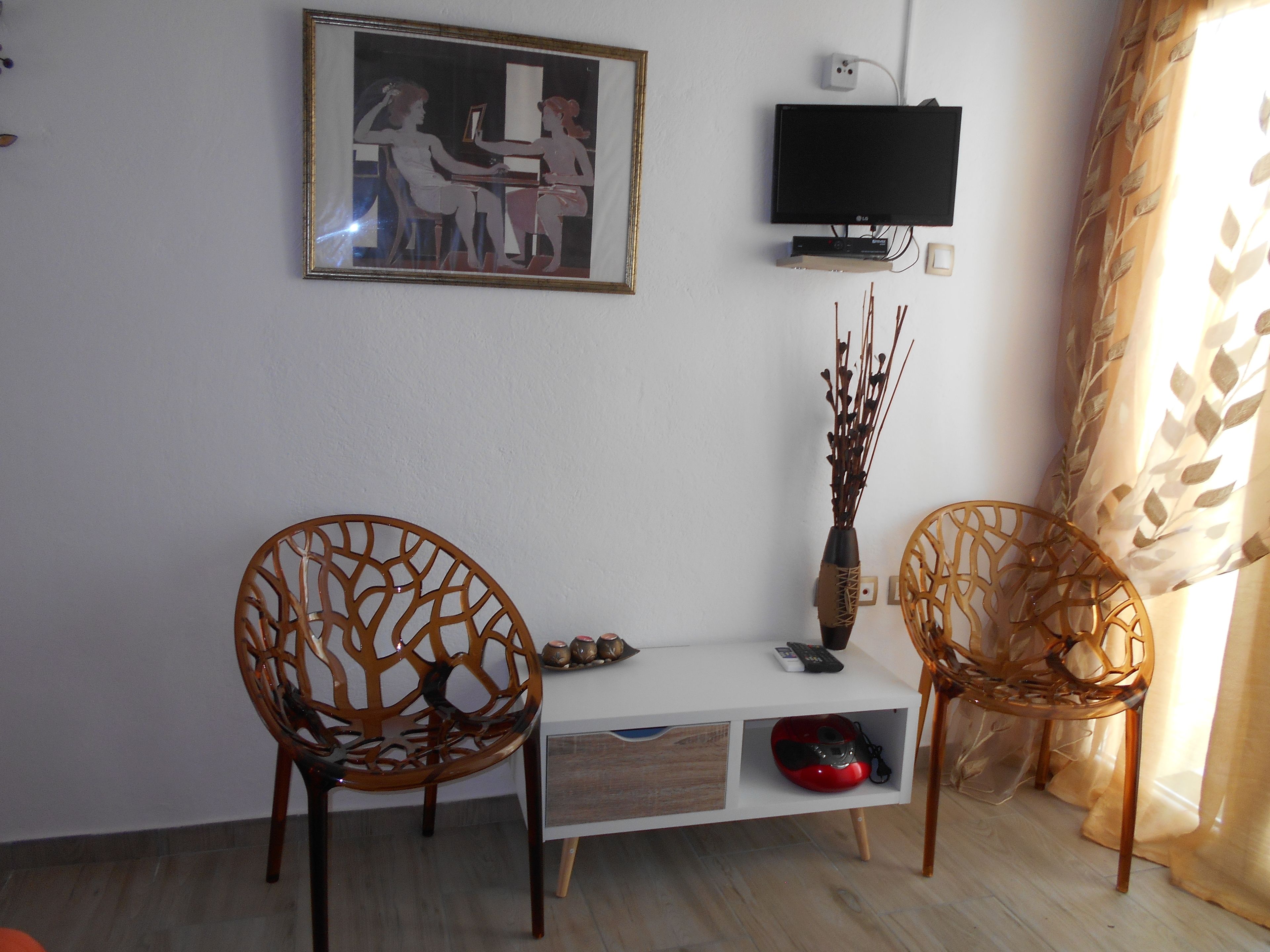 Sala de estar com duas cadeiras de madeira, um rack branco, um quadro emoldurado, uma TV de tela plana e um vaso com galhos.