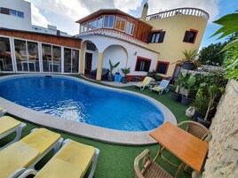 Piscina com cadeiras de descanso amarelas e uma mesa de madeira, cercada por plantas em vasos e um edifício com sacadas.