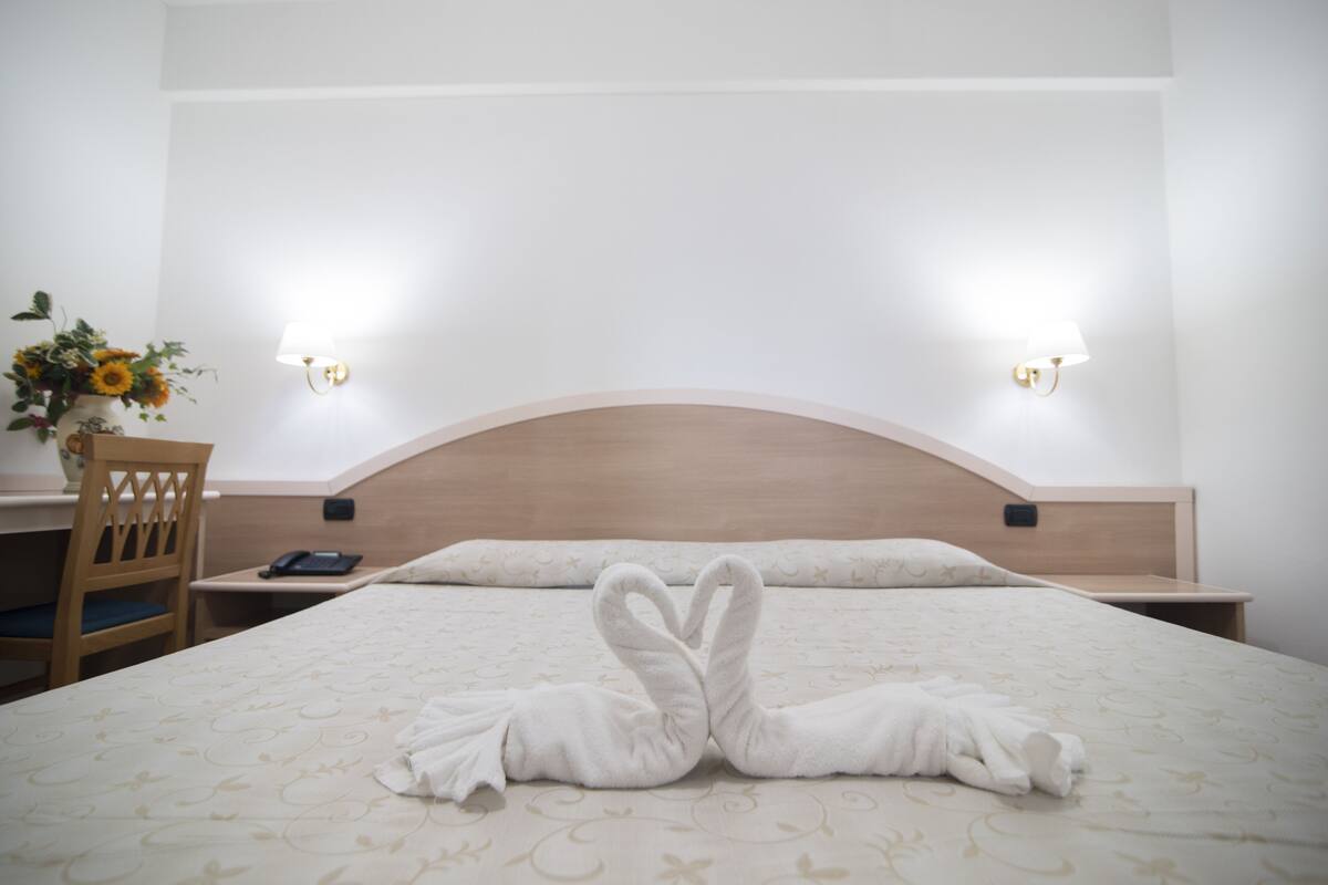 Quarto de hotel com cama, duas mesas de cabeceira, uma poltrona, um vaso com flores e luminárias na parede.