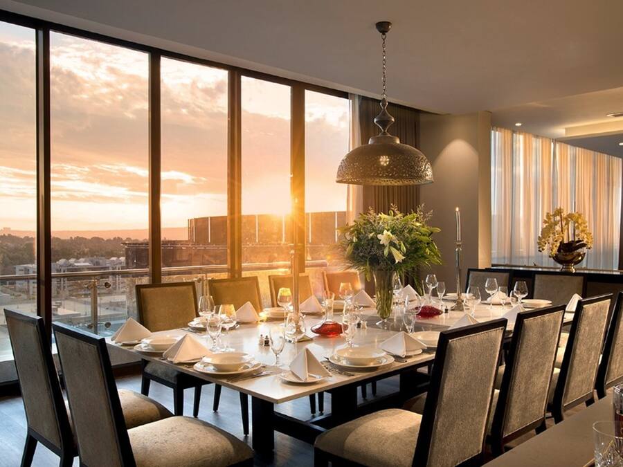 Uma sala de jantar com mesa posta para seis pessoas, janelas amplas com vista para a cidade e um lustre.