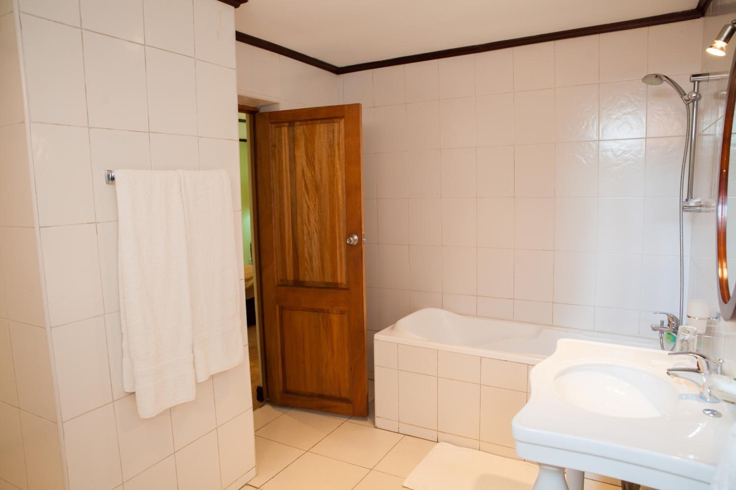 Um banheiro com porta de madeira, paredes com azulejos brancos, uma banheira branca e uma pia com espelho.